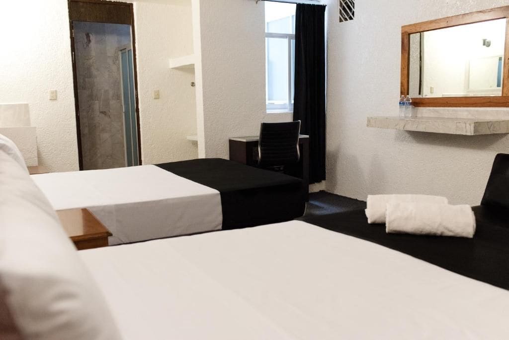 Quarto de hotel com duas camas, uma escrivaninha, uma cadeira, um espelho e uma janela com cortinas.