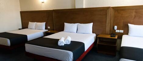 Quarto de hotel com duas camas, cada uma com lençóis brancos e um toque de vermelho. Há um ventilador de teto, uma mesa de cabeceira com uma luminária e uma luminária fixada na parede.