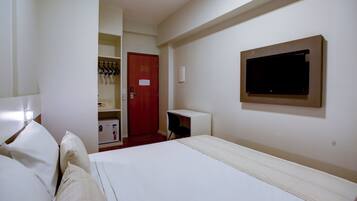 Quarto de hotel com uma cama, uma escrivaninha, uma televisão e uma porta que dá para um banheiro.