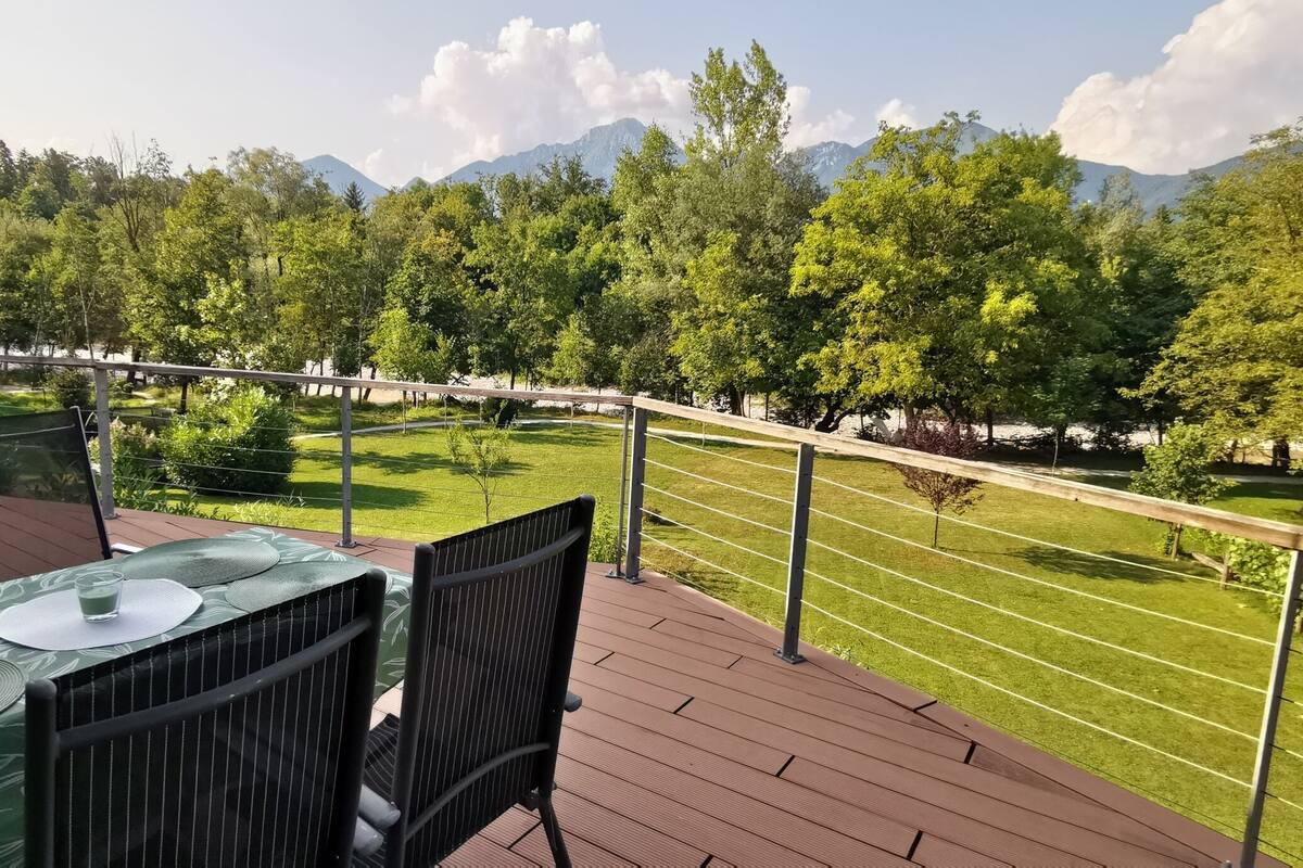 Um terraço com mesa e cadeiras, com vista para um gramado bem cuidado e uma área arborizada com montanhas ao fundo.