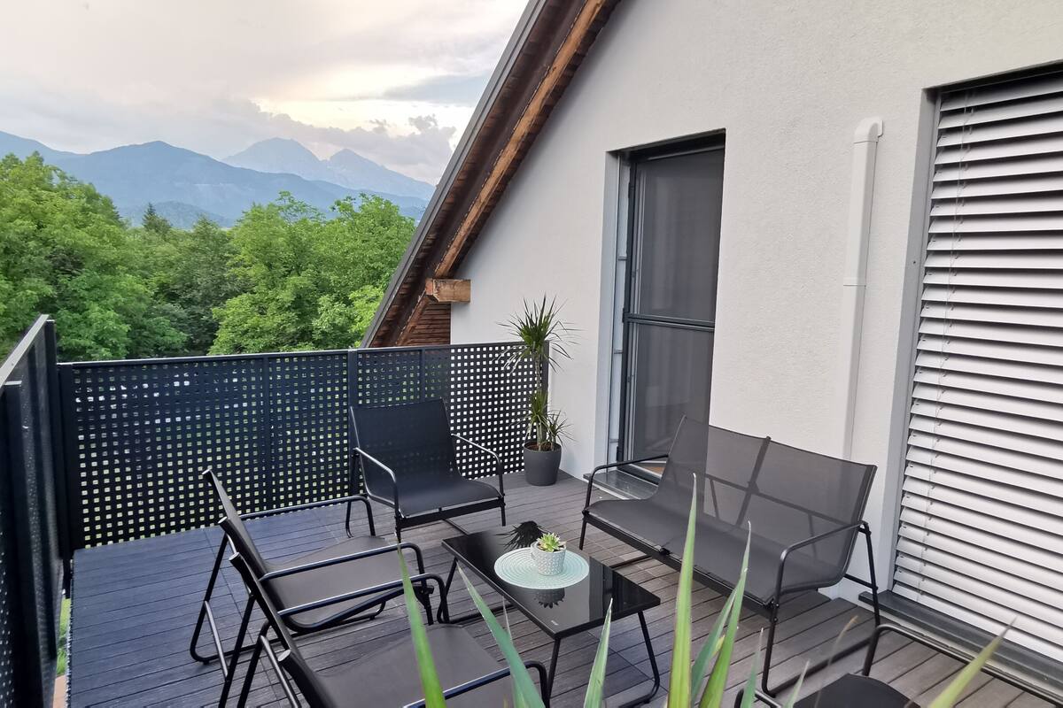 Uma varanda com vista para montanhas, uma cadeira de metal preta, uma pequena mesa com uma planta e uma porta deslizante fechada.