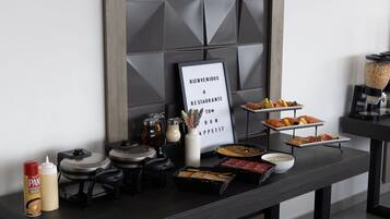 Disposição de buffet com placa de boas-vindas, diversos alimentos, acompanhamentos e uma máquina de café.