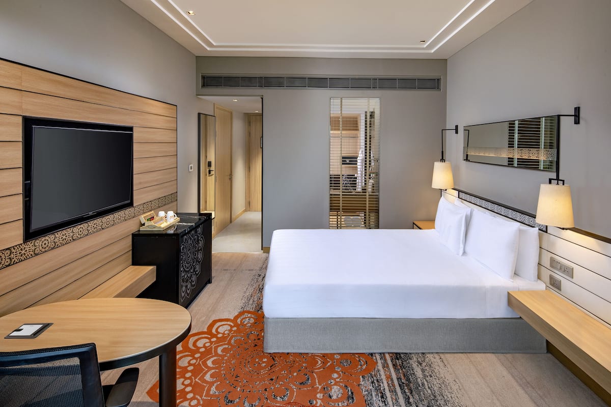Quarto de hotel moderno com uma cama grande, televisão na parede, escrivaninha com cadeira e uma mesinha.