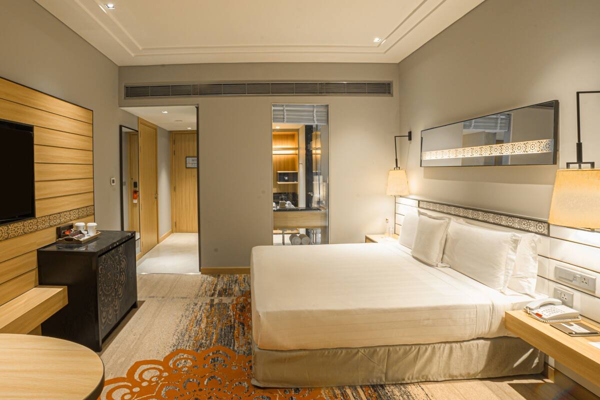 Quarto de hotel com uma cama, uma escrivaninha, uma televisão, um espelho e um banheiro visível por uma porta aberta.