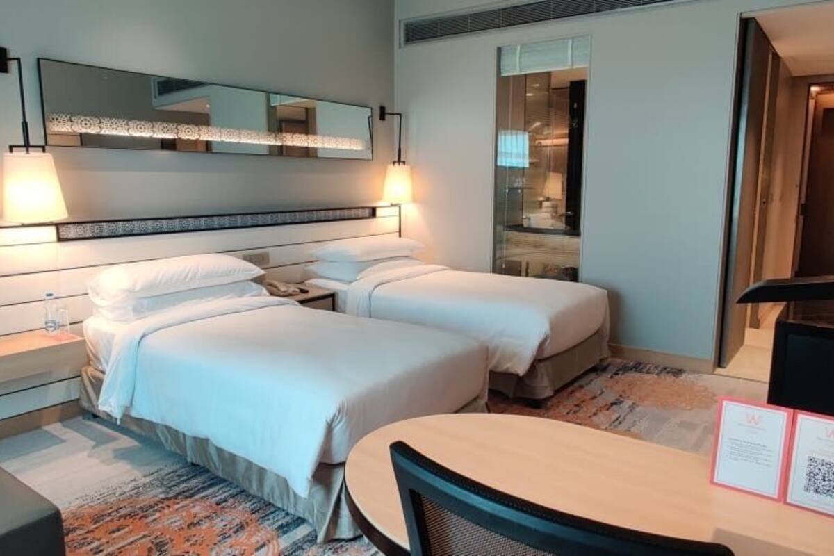 Quarto de hotel com duas camas, uma escrivaninha, uma cadeira, um espelho e uma pequena mesa com um código QR.