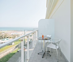 Varanda com guarda-corpo de vidro com vista para uma praia, uma mesa pequena com cadeiras e uma bebida.