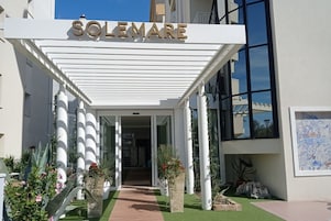 Entrada de hotel com toldo branco e colunas, céu claro e placa com a inscrição 'SOLEMARE'.