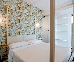 Quarto com cama, mesa de cabeceira, parede com papel de parede tropical e porta que dá para um armário.