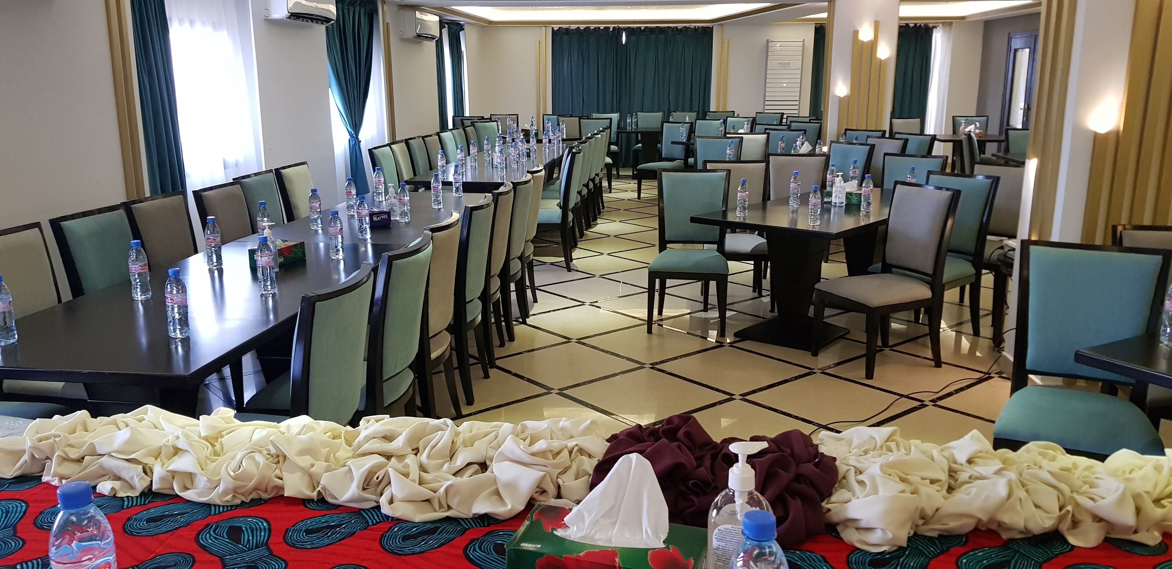 Uma sala de conferências de hotel com várias mesas e cadeiras, garrafas de água sobre as mesas e uma pilha de roupas amassadas no chão.