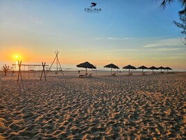 Uma praia com espreguiçadeiras e guarda-sóis de palha, baloiço e céu claro.
