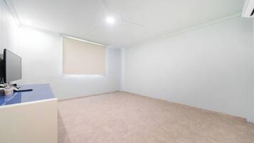 Quarto com escrivaninha, televisão, janela com persianas e piso coberto por carpete.