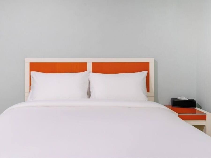 Uma cama com roupa de cama branca e cabeceira laranja, uma mesa de cabeceira com um objeto preto, e uma parede lisa.