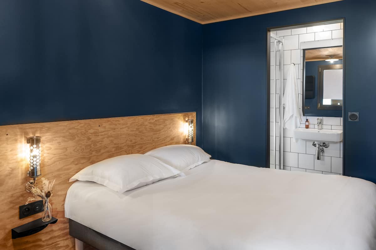 Quarto com cabeceira de madeira, cama branca, mesa de cabeceira com abajur e banheiro com espelho e pia.