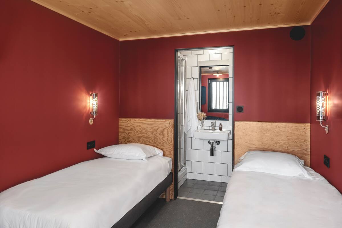 Quarto de hotel com duas camas de solteiro, teto de madeira, paredes vermelhas e banheiro com espelho e pia.