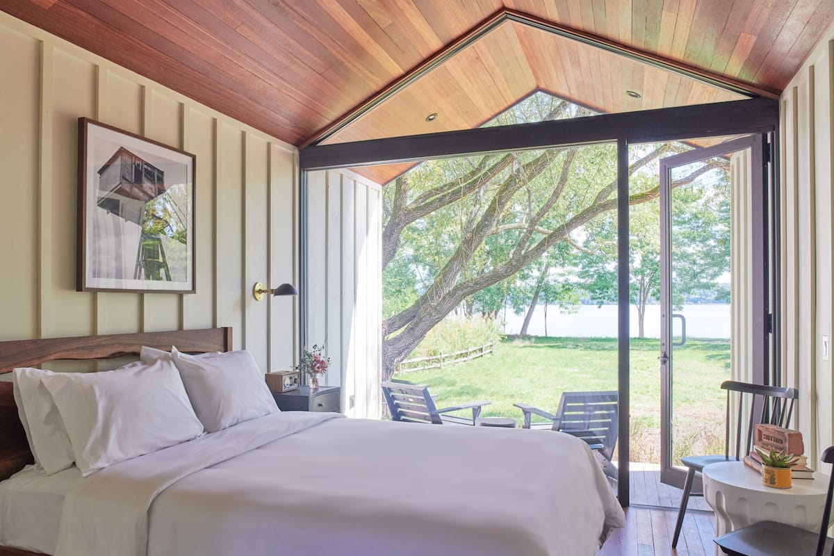 Quarto com uma cama grande, teto de madeira e vista para uma área gramada através de portas de vidro.