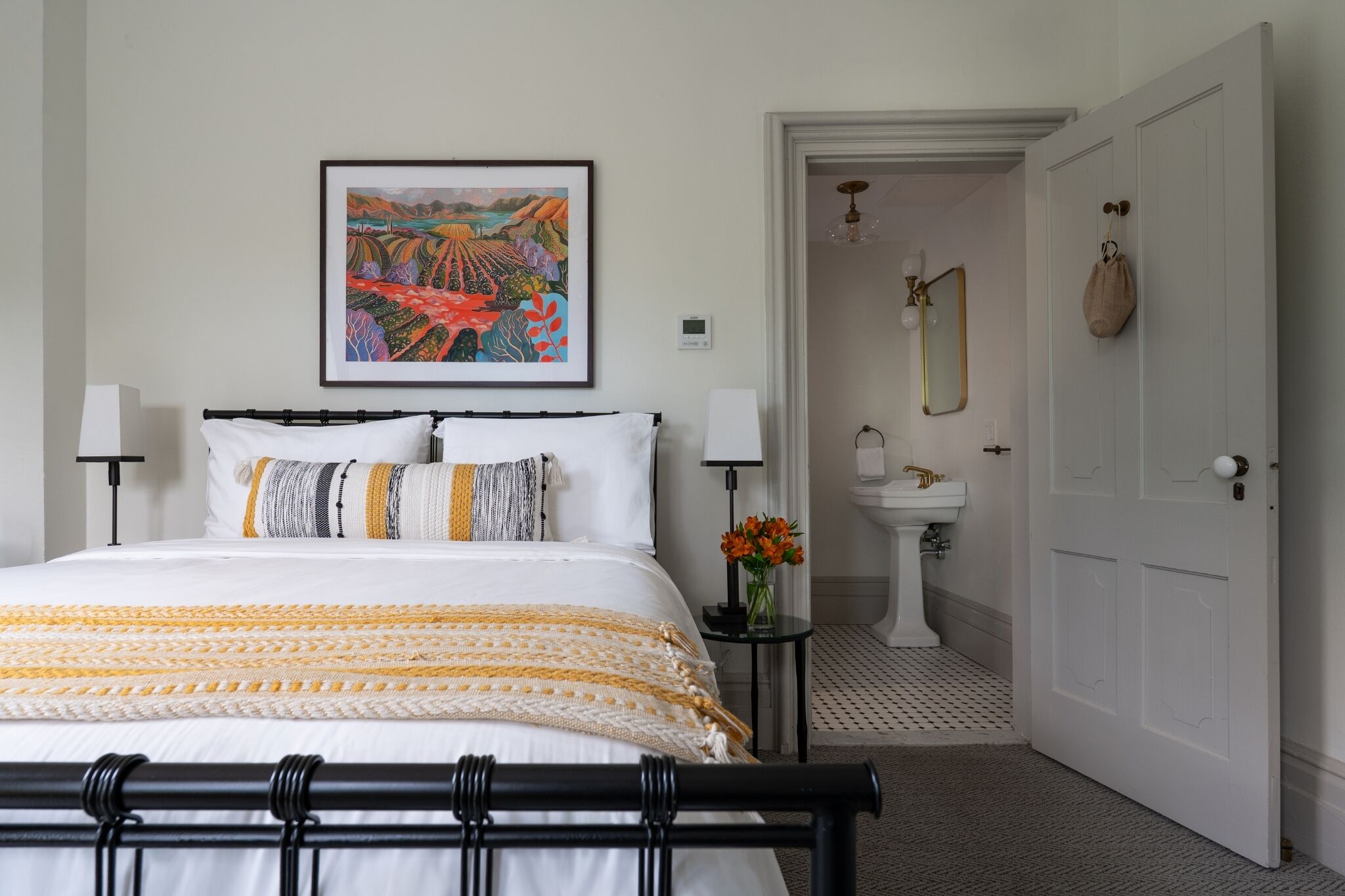 Quarto de hotel com cama, mesa de cabeceira com luminária, um quadro na parede e banheiro visível por uma porta aberta.