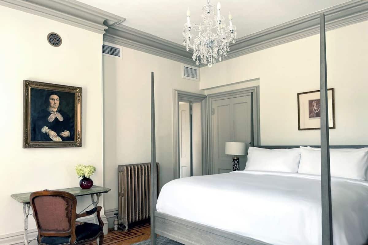 Quarto de hotel com uma cama grande, uma escrivaninha com cadeira, um candelabro e obras de arte emolduradas nas paredes.