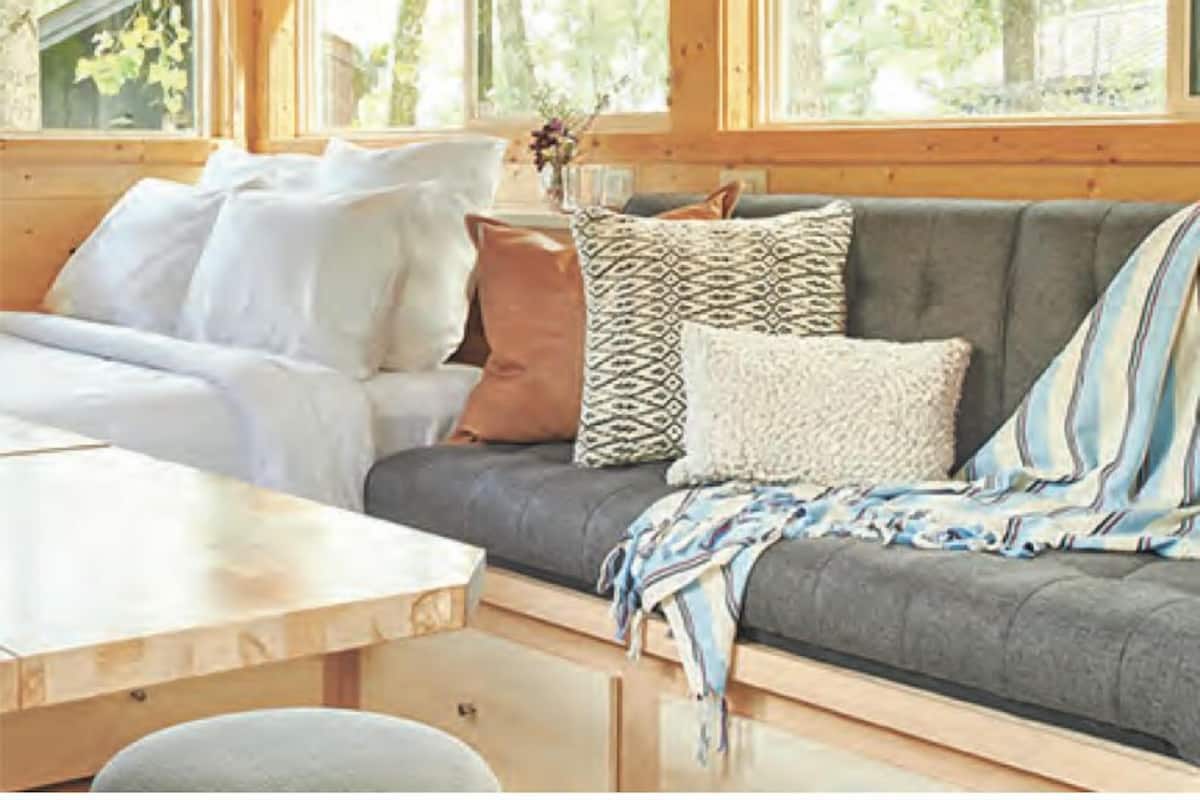 Interior acolhedor de cabana com teto de madeira, um sofá com almofadas, uma mesa e janelas amplas que oferecem vista para o exterior.