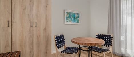 Quarto com guarda-roupa de madeira, duas cadeiras com assento trançado, uma mesa de madeira redonda e um quadro abstrato emoldurado na parede.