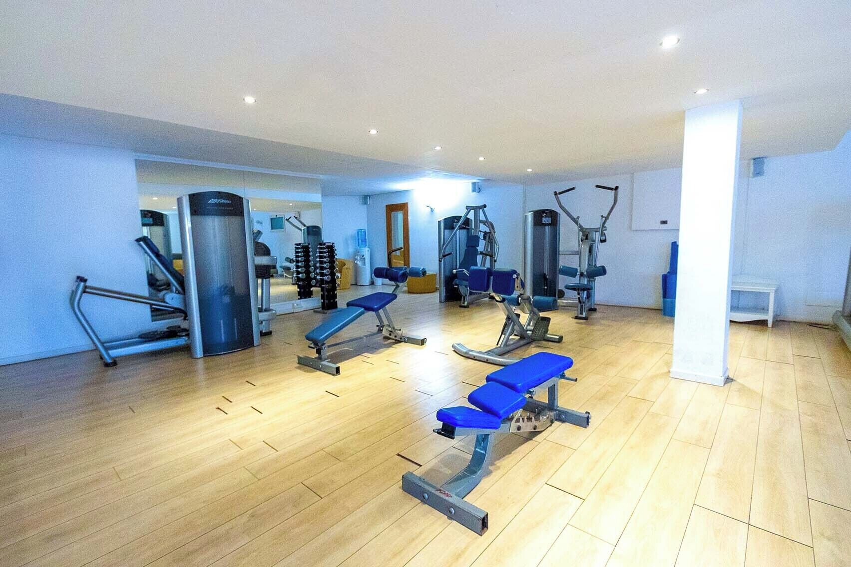 Uma academia com diversos equipamentos de exercício, incluindo bancos e máquinas de peso, e um painel de espelhos.