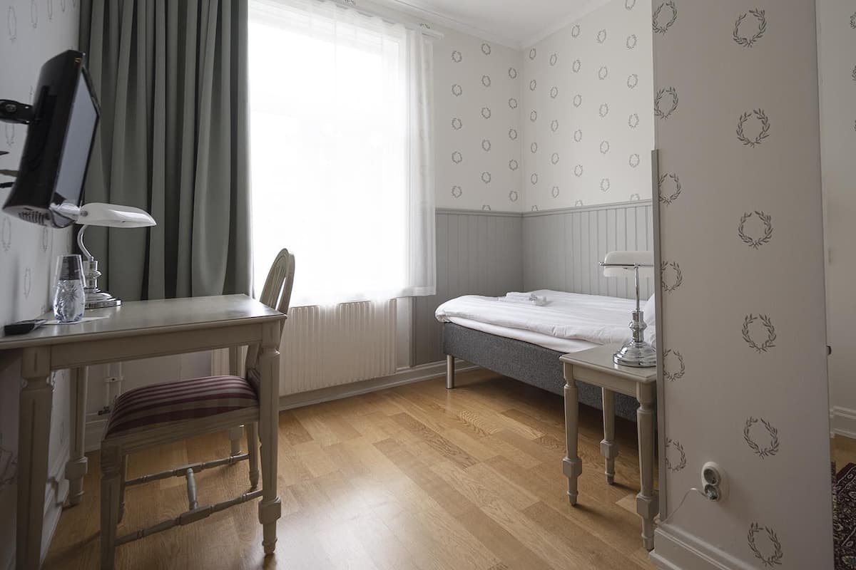 Quarto com uma cama de solteiro, mesa de cabeceira, cadeira, escrivaninha com abajur e janela com cortinas.