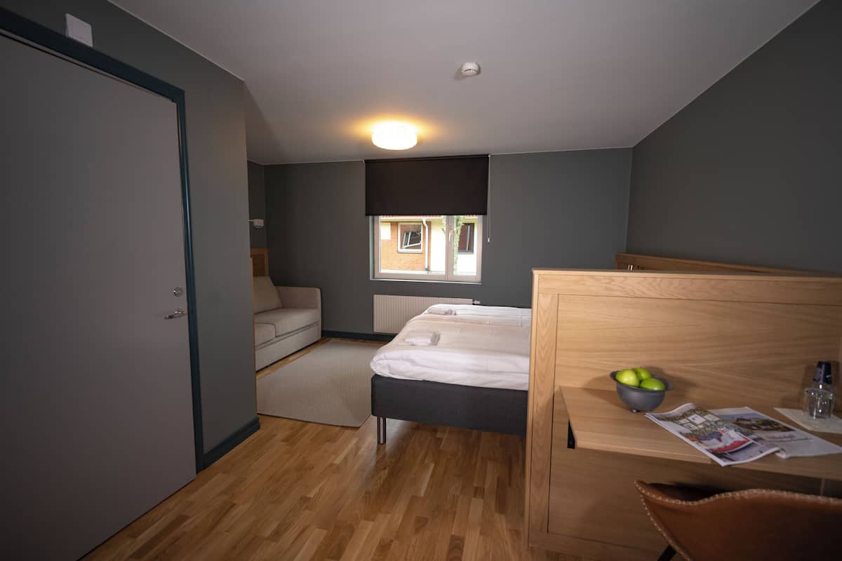 Um quarto com uma cama, uma escrivaninha com uma tigela de fruta, uma cadeira, uma janela e uma porta.
