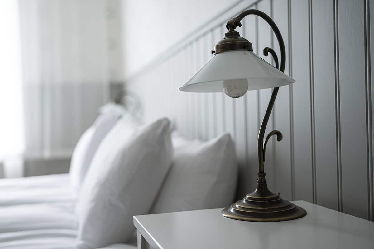 Uma mesinha de cabeceira com abajur, uma cama com roupa de cama branca e uma janela com cortinas.