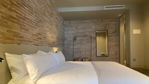 Quarto de hotel com parede de tijolos, uma cama grande com roupa de cama branca, uma mesinha de cabeceira, um espelho e uma porta que dá para outro quarto.