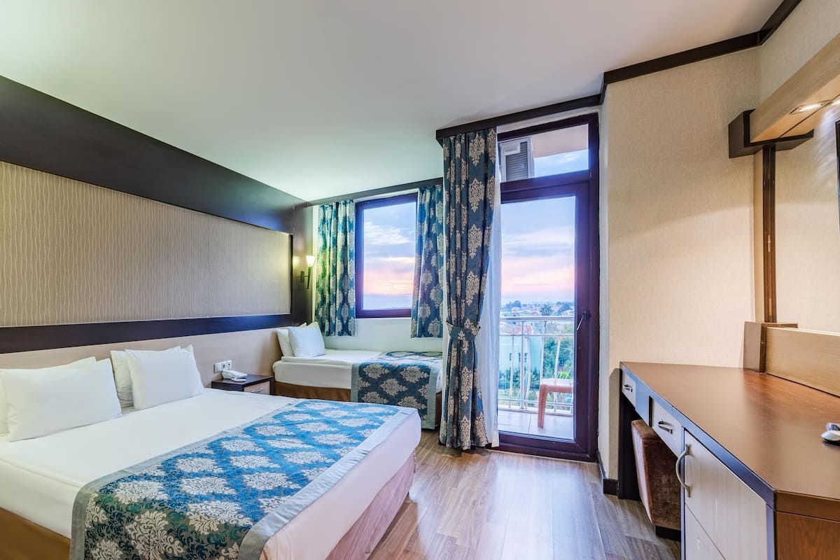 Quarto de hotel com cama grande, escrivaninha, televisão e varanda com cortinas.