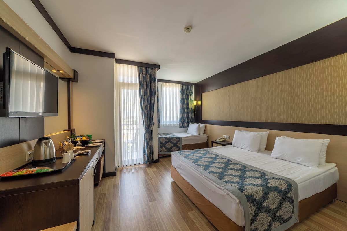 Quarto de hotel com uma cama grande, uma escrivaninha com chaleira, televisão e janela com cortinas.
