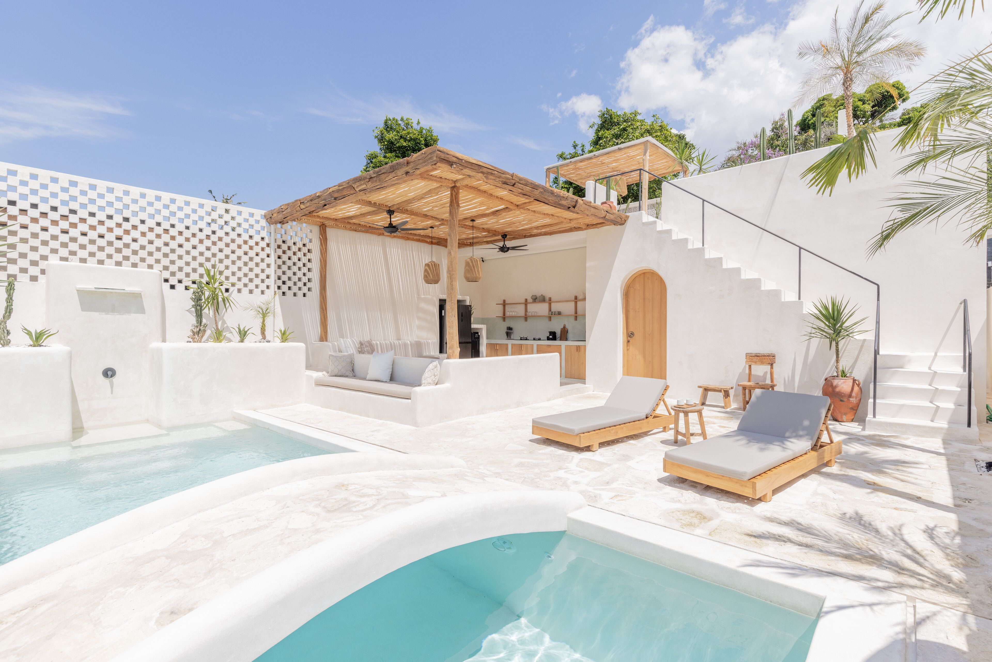 Área de piscina com espreguiçadeiras, uma pérgola e uma escadaria que leva a uma cozinha ao ar livre coberta.
