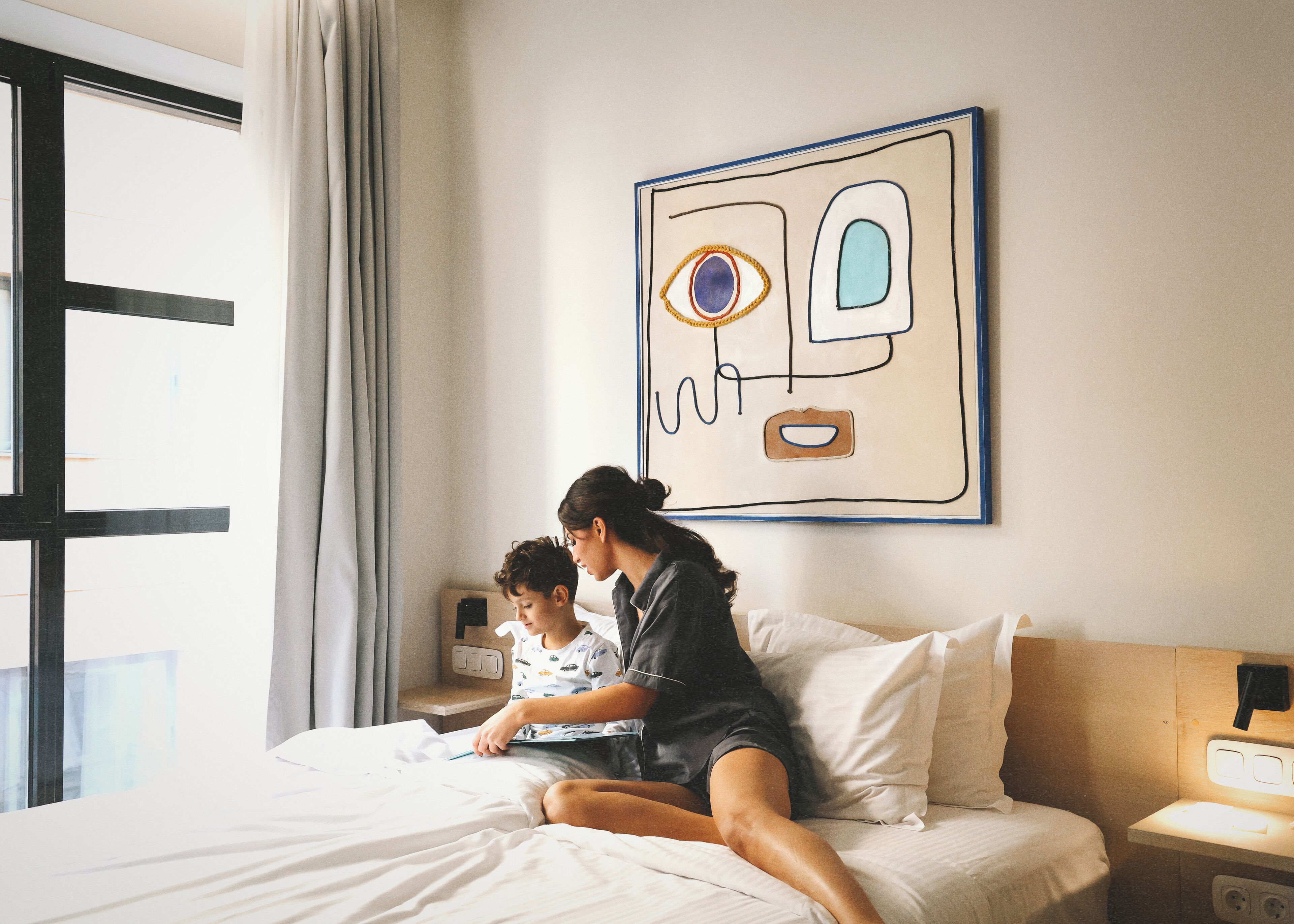 Quarto de hotel com uma cama, mesa de cabeceira, luminária na parede e janela com cortinas.