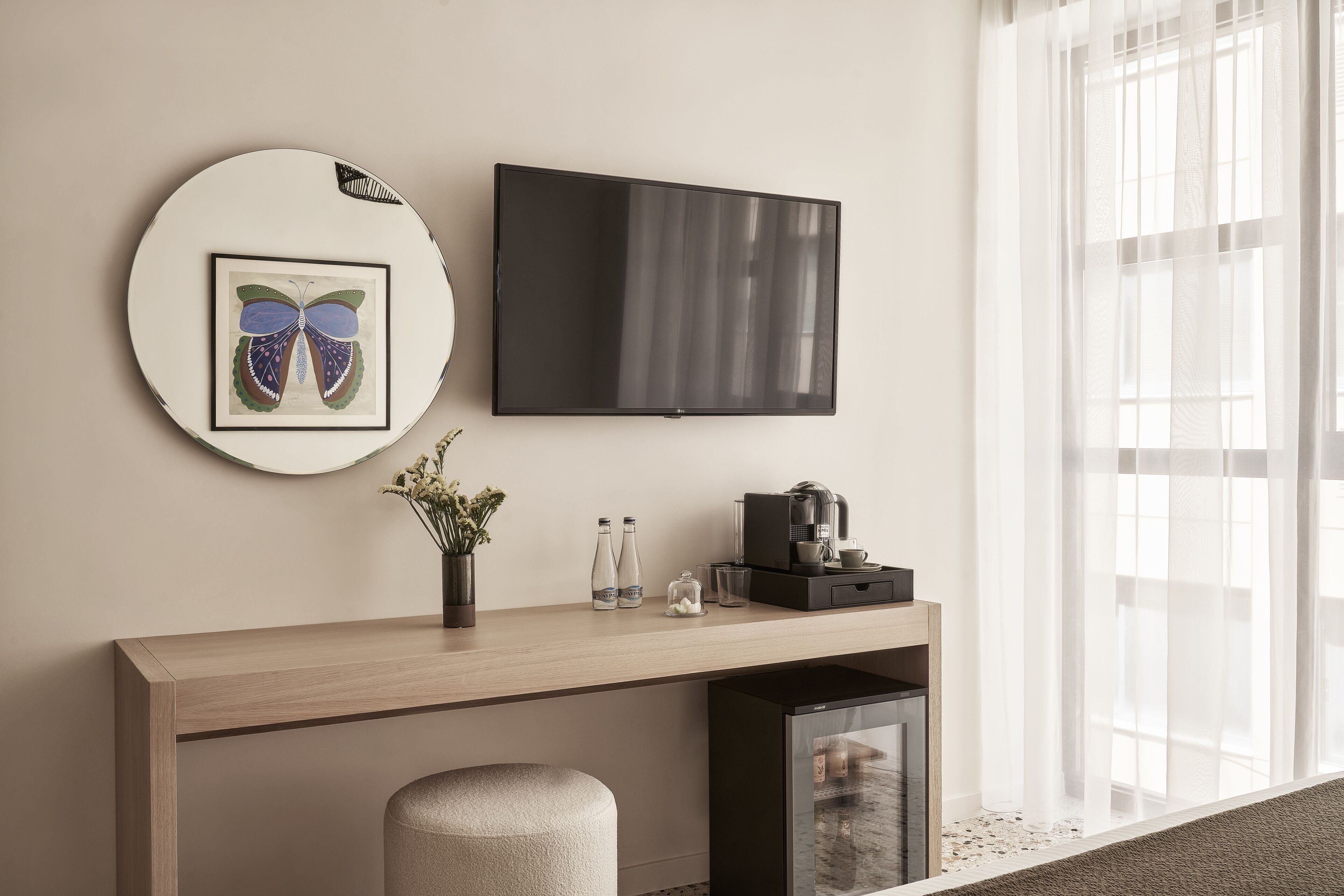 Quarto de hotel moderno com televisão de tela plana na parede, mesinha de madeira com cafeteira e um pequeno espelho redondo.