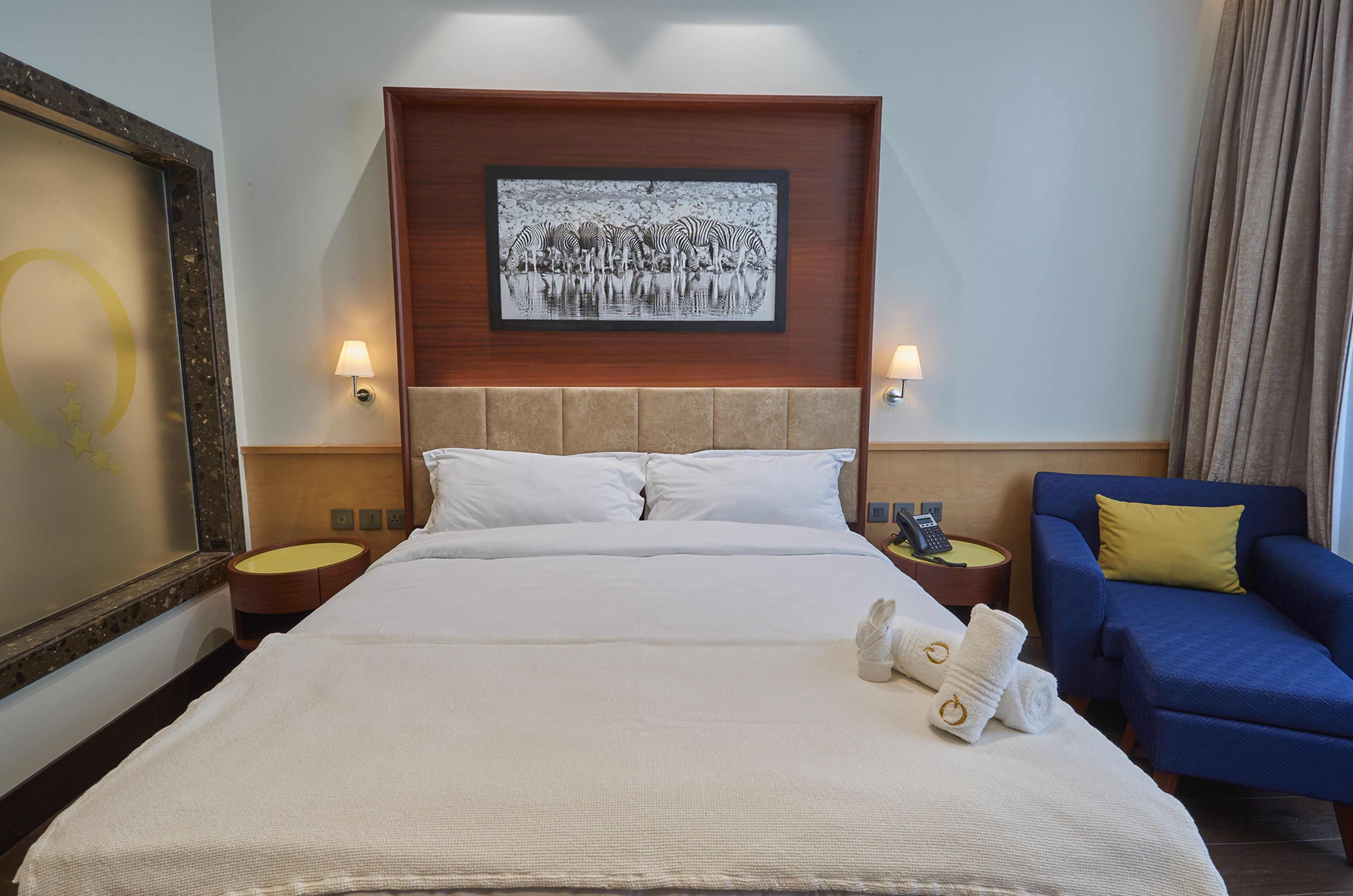Quarto de hotel com uma cama grande, cabeceira com quadro emoldurado, duas luminárias de cabeceira, uma poltrona azul e uma mesinha com telefone e abajur.