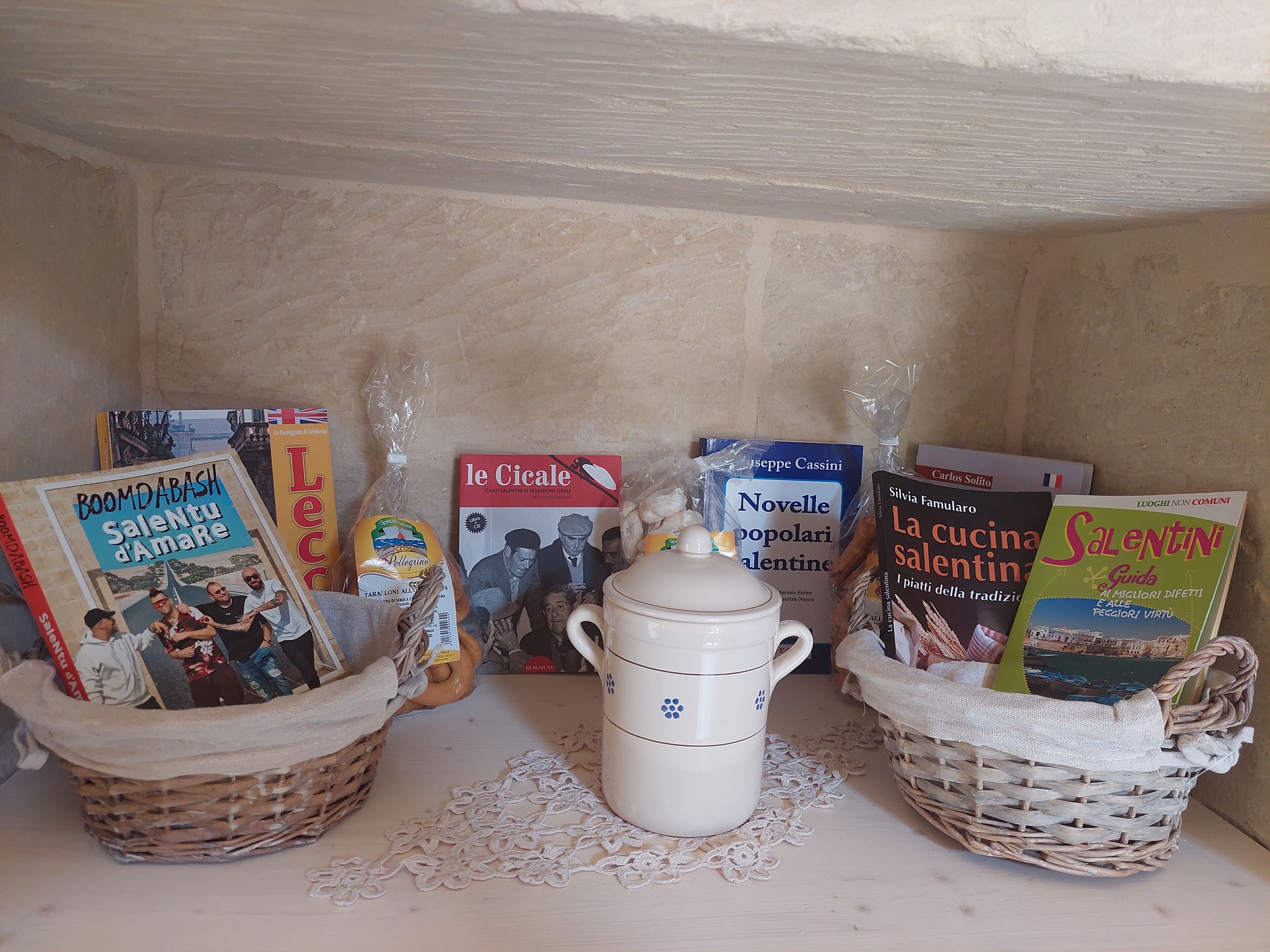 Uma bancada de cozinha com cestos contendo livros, uma garrafa de azeite e um jarro de cerâmica branca.