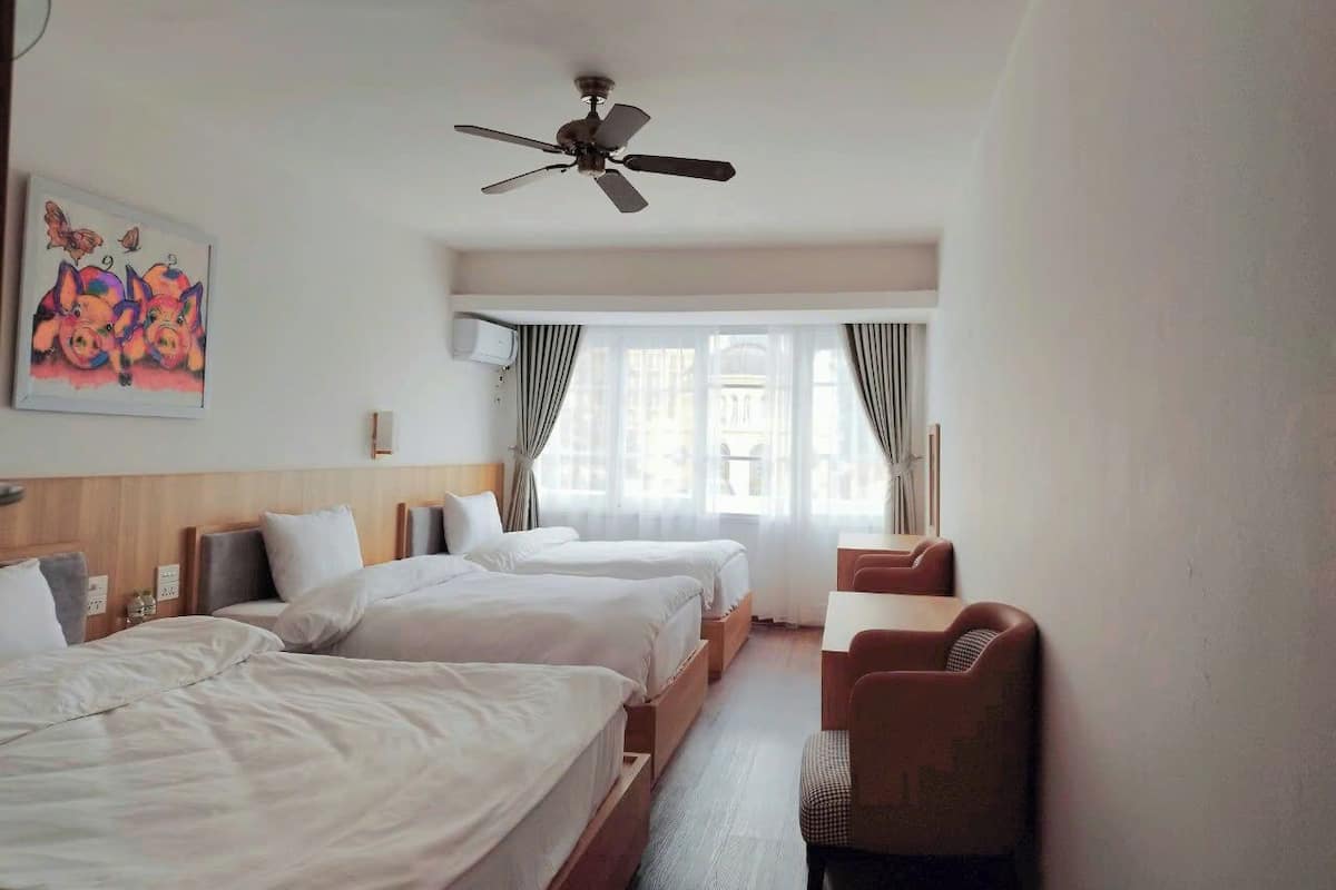 Quarto de hotel com duas camas, um ventilador de teto, uma janela com cortinas e uma mesa com cadeira.