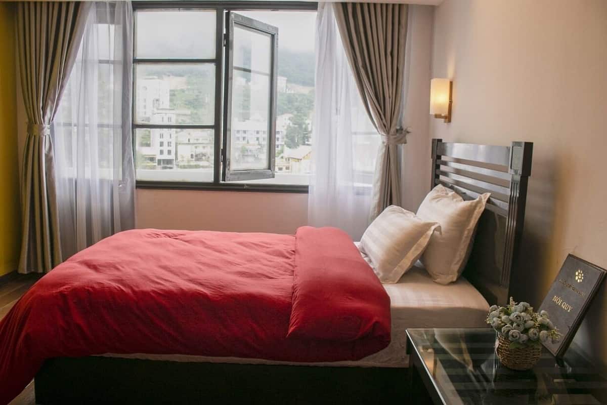 Quarto de hotel com cama, mesa de cabeceira, janela com cortinas e vista para edifícios.
