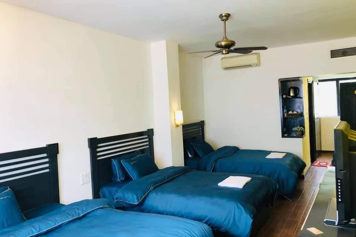 Quarto de hotel com duas camas, um ventilador de teto, um ar condicionado fixado na parede e uma pequena estante com objetos.