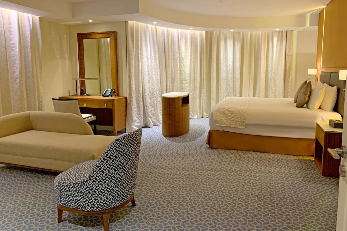 Quarto de hotel com cama, escrivaninha com telefone, cadeira e um banquinho redondo.