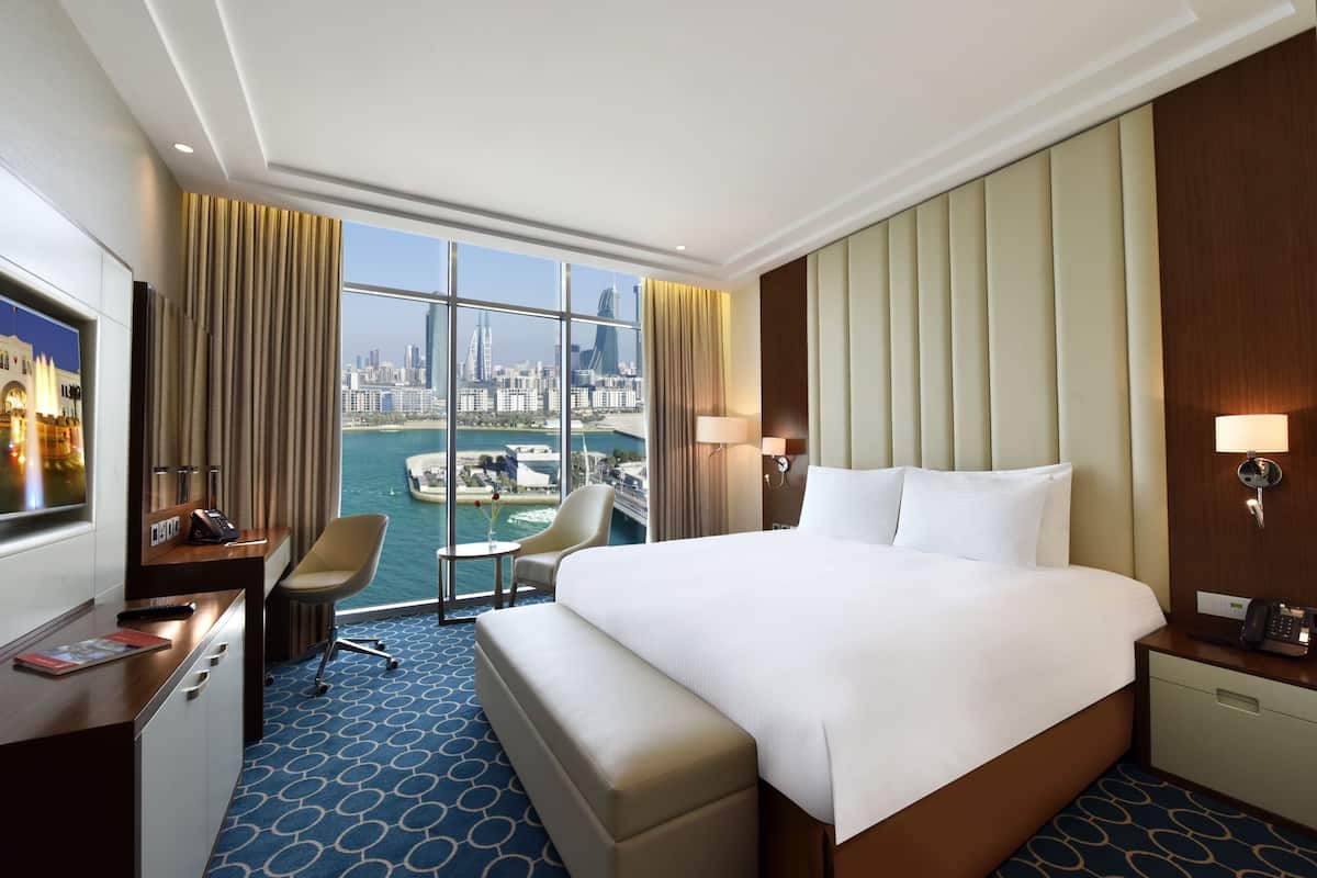 Quarto de hotel com uma cama grande, uma escrivaninha, uma cadeira e vista para o horizonte da cidade.