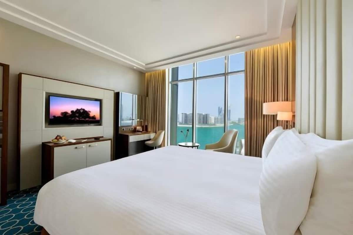 Quarto de hotel com uma cama grande, uma escrivaninha com cadeira, televisão e janela com vista para a cidade.