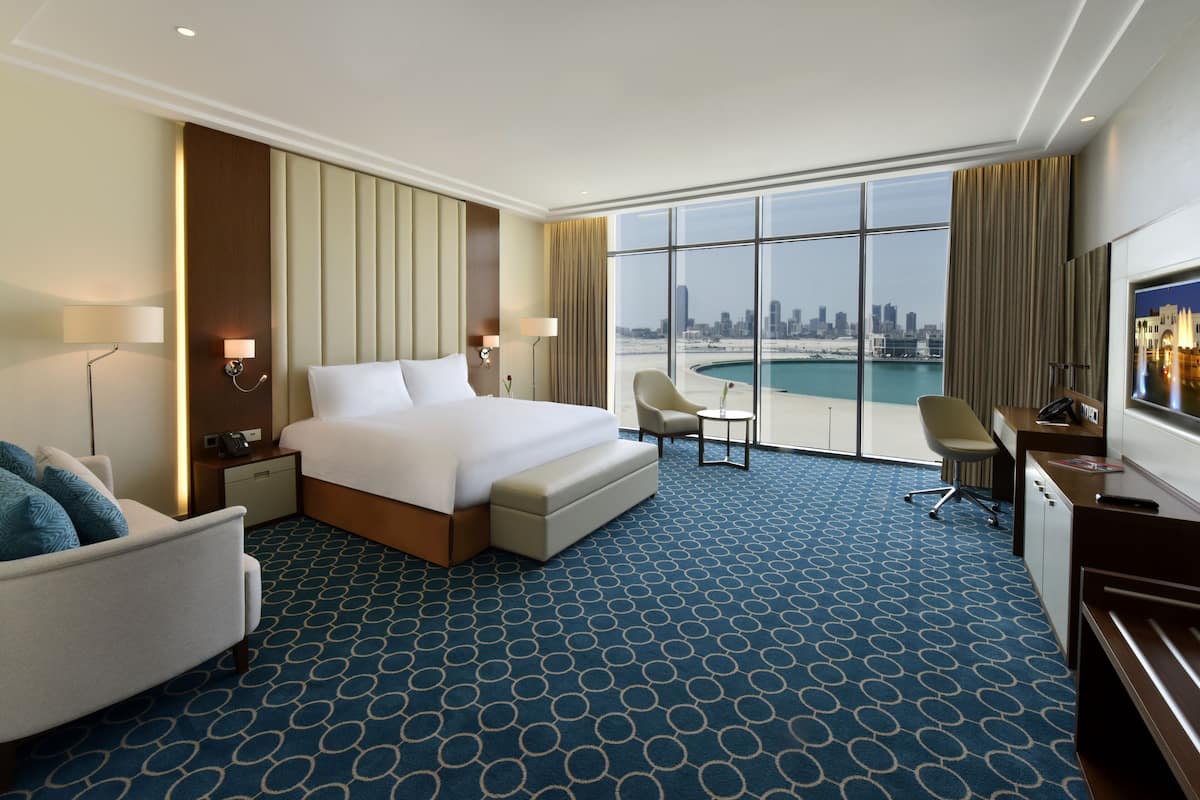 Quarto de hotel com uma cama grande, uma escrivaninha, uma cadeira e vista para o horizonte urbano e para a água.