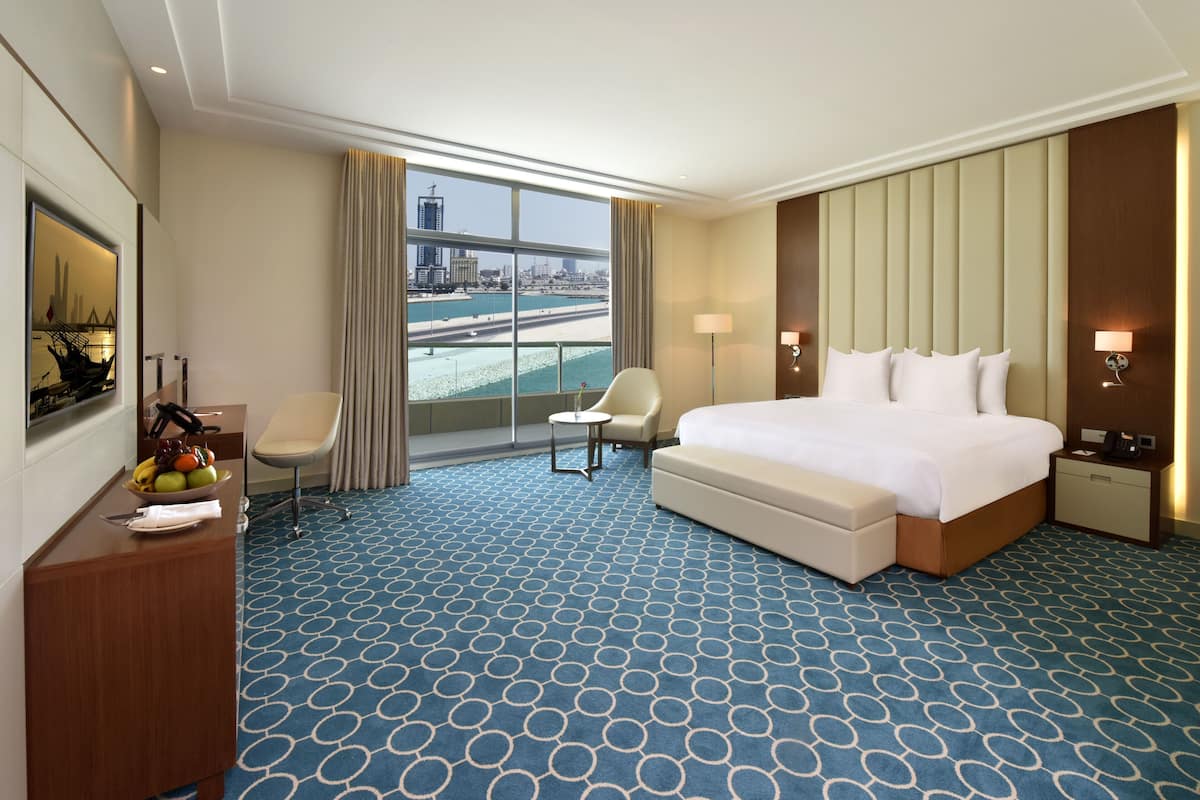 Quarto de hotel com uma cama grande, uma escrivaninha com uma fruteira, uma cadeira, uma mesa de cabeceira e vista para a paisagem urbana através de grandes janelas.