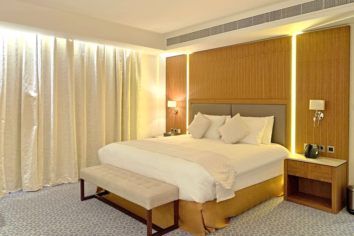 Quarto de hotel com uma cama grande, mesas de cabeceira, um banquinho e cortinas.