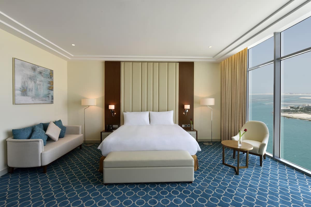 Quarto de hotel com uma cama grande, um sofá, uma poltrona e vista para o mar.