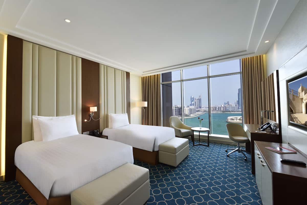 Quarto de hotel com duas camas, uma escrivaninha, uma cadeira e vista para a cidade.