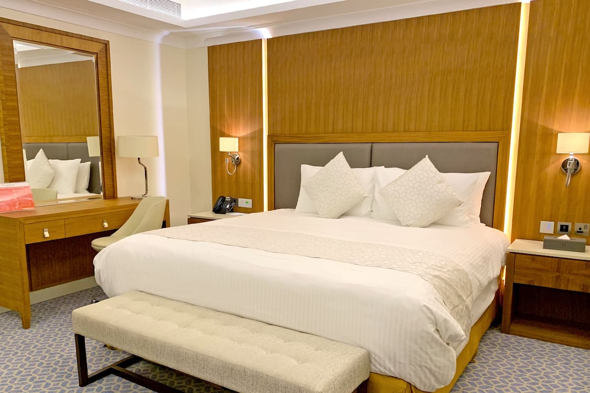 Quarto de hotel com uma cama grande, mesa de cabeceira com abajur, escrivaninha com cadeira e um espelho.