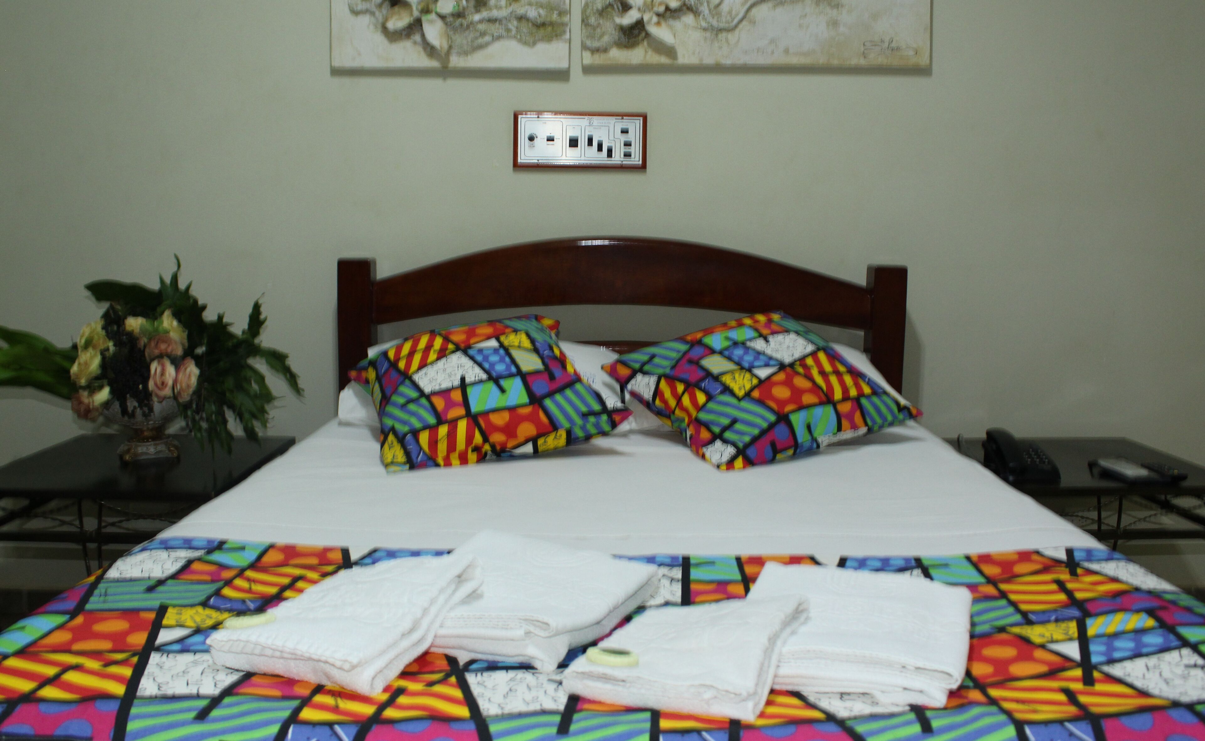 Uma cama com almofadas coloridas e um edredom, uma mesa de cabeceira com telefone e controles remotos, e uma tomada na parede acima da cama.