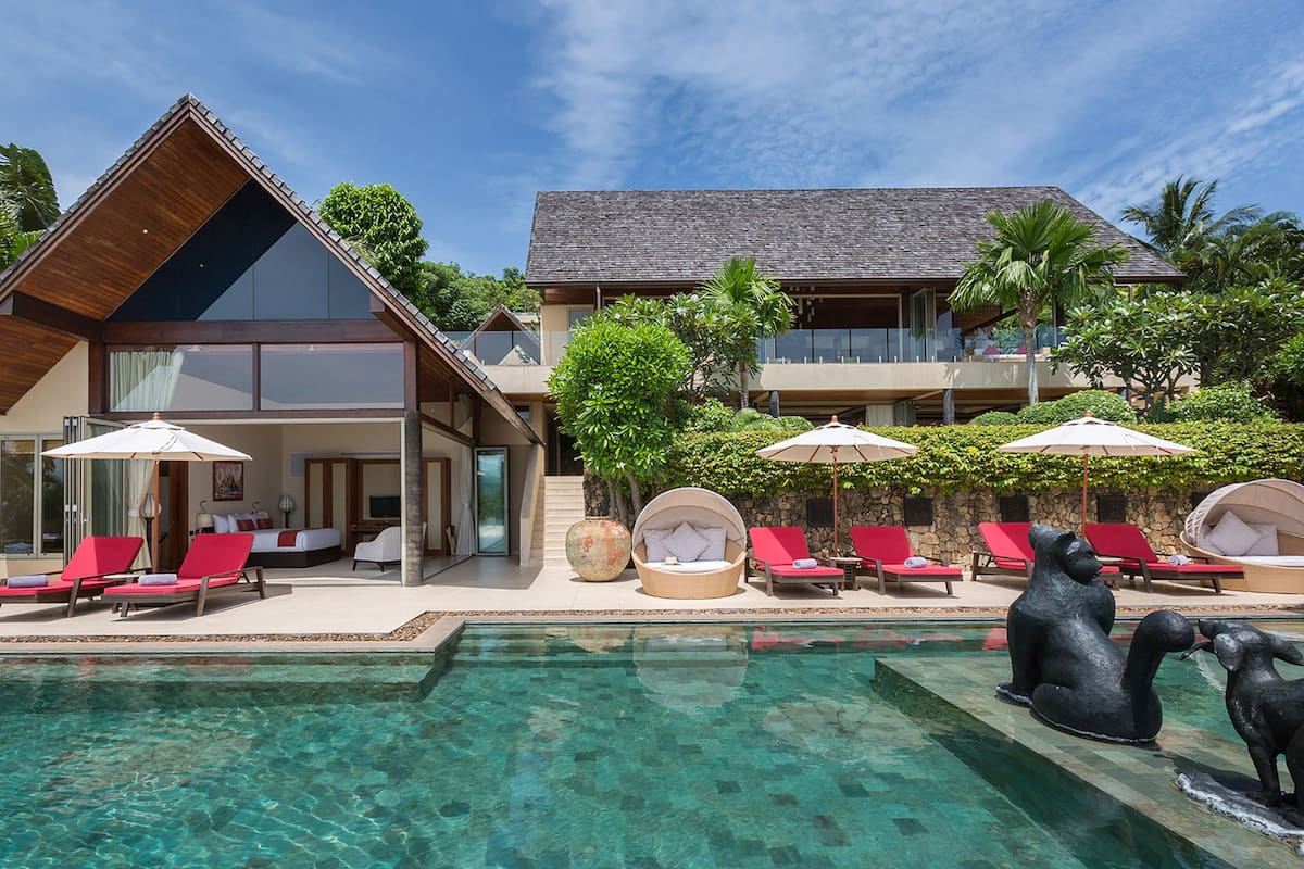 Um resort com piscina transparente, cadeiras de sol vermelhas e um edifício grande com varandas.