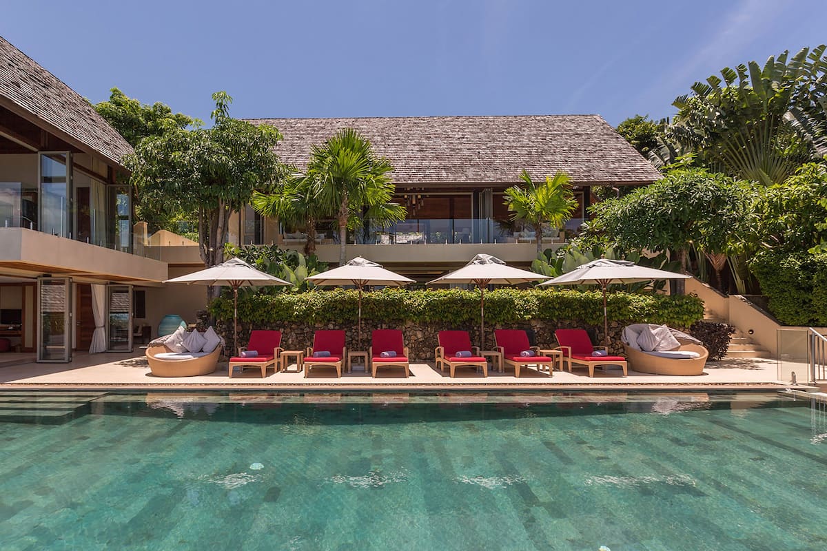 Uma área de piscina com espreguiçadeiras e guarda-sóis, piscina translúcida e um edifício com telhado de azulejos e vegetação.
